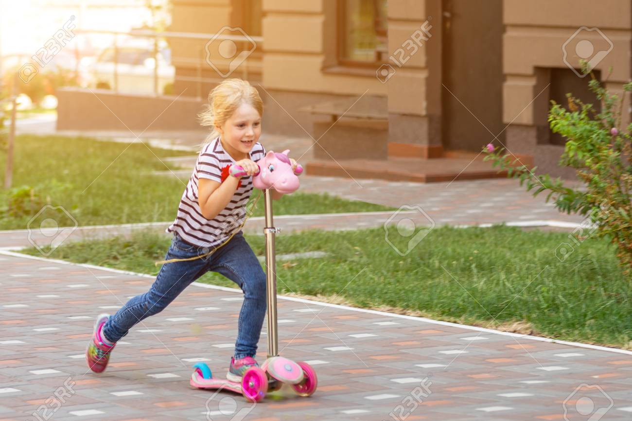 Cute Little Girl Riding Scooter In City Park On Bright Summer Day. Blond Toddler Having Fun On Street. Active Leisure And Outdoor Sport For Children Concept Stock Photo Picture and Royalty Free